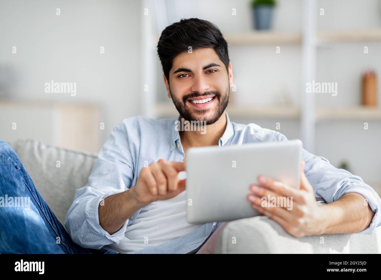 Portrait of excited arab man using tablet, sitting on couch at home and ...
