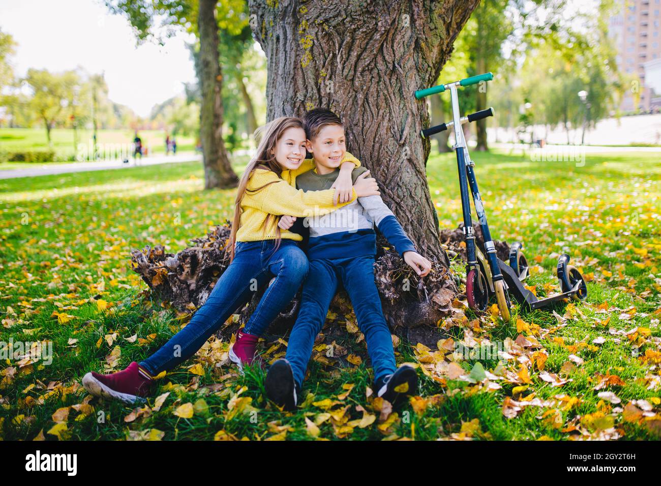 Friends kids posing happily sitting under a tree in an autumn park next ...