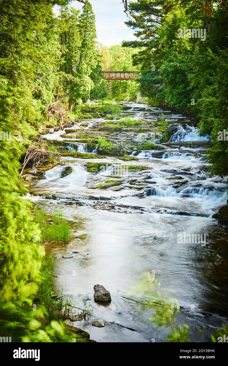 View through trees of waterfalls cascading down river with bridge in ...