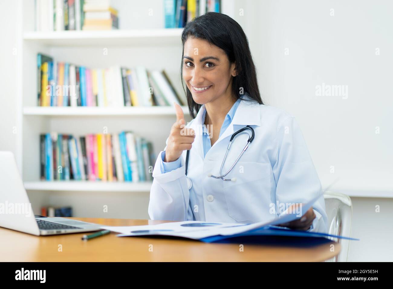 Laughing latin american female gynecologist at desk at work at hospital ...