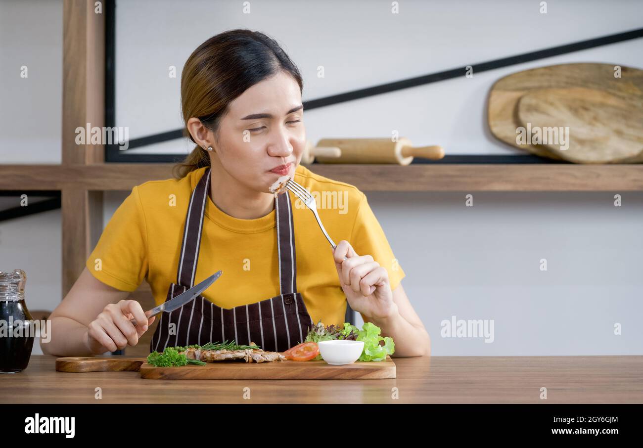 Young asian woman in brown apron enjoy eating the steak while closing ...