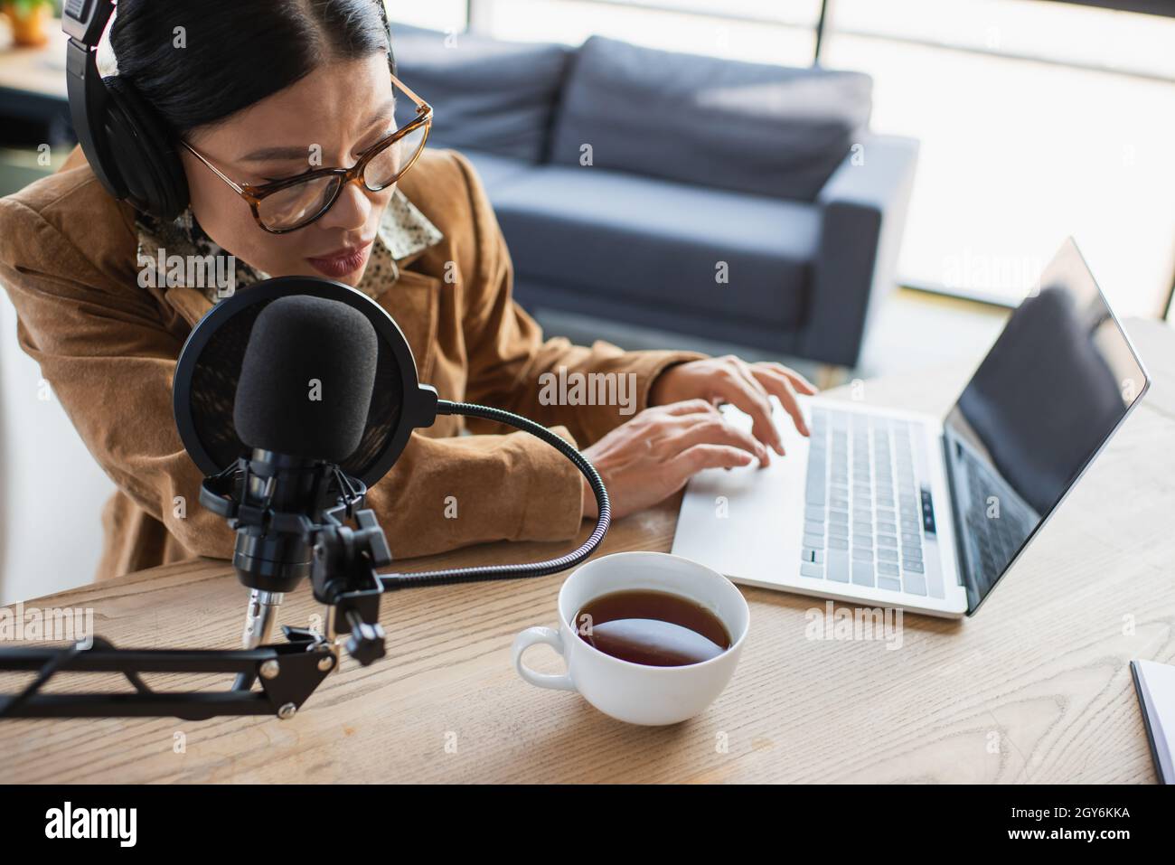 asian radio host in glasses talking in microphone while using laptop in ...