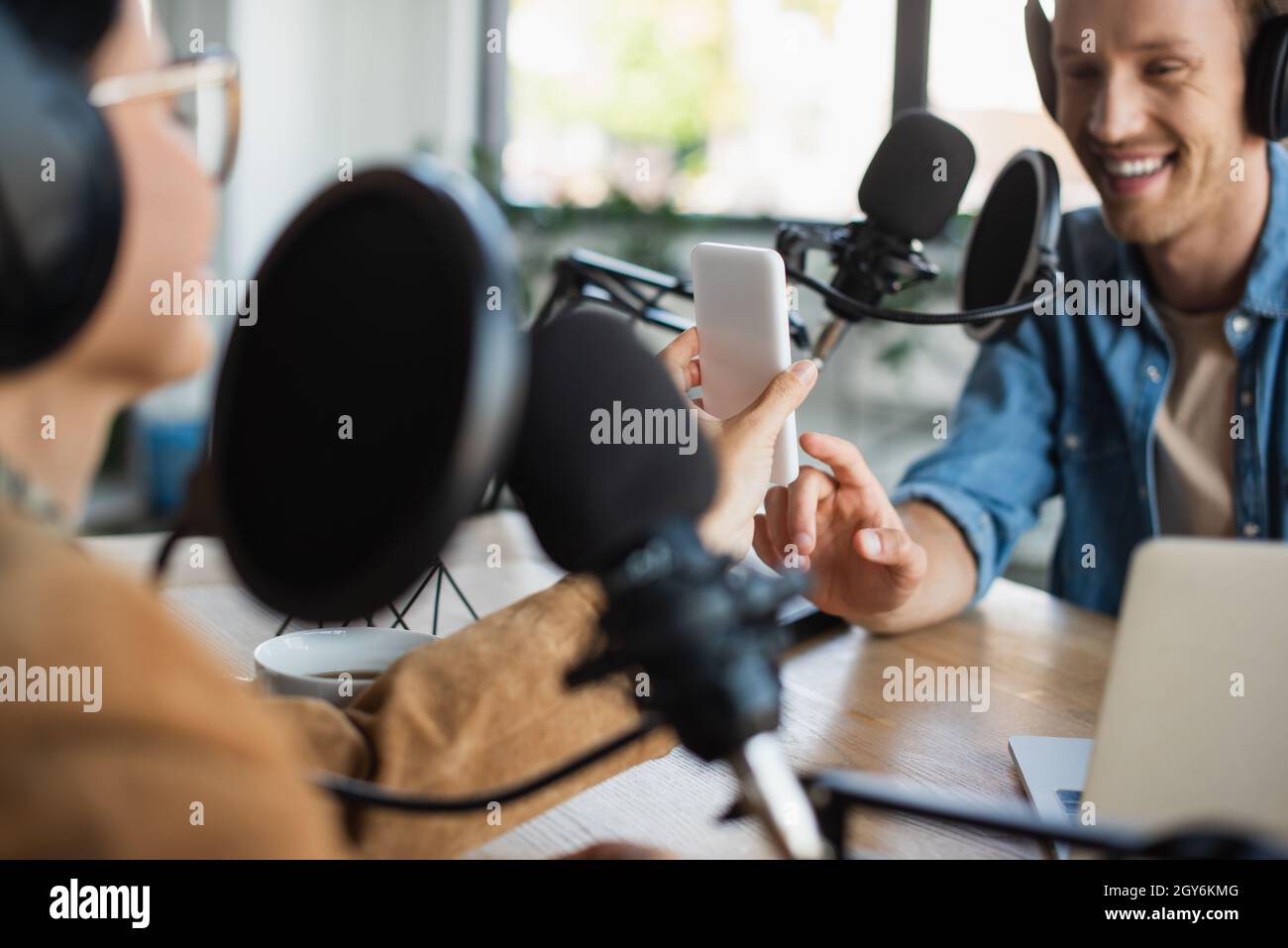 happy radio host pointing at smartphone near blurred asian colleague ...