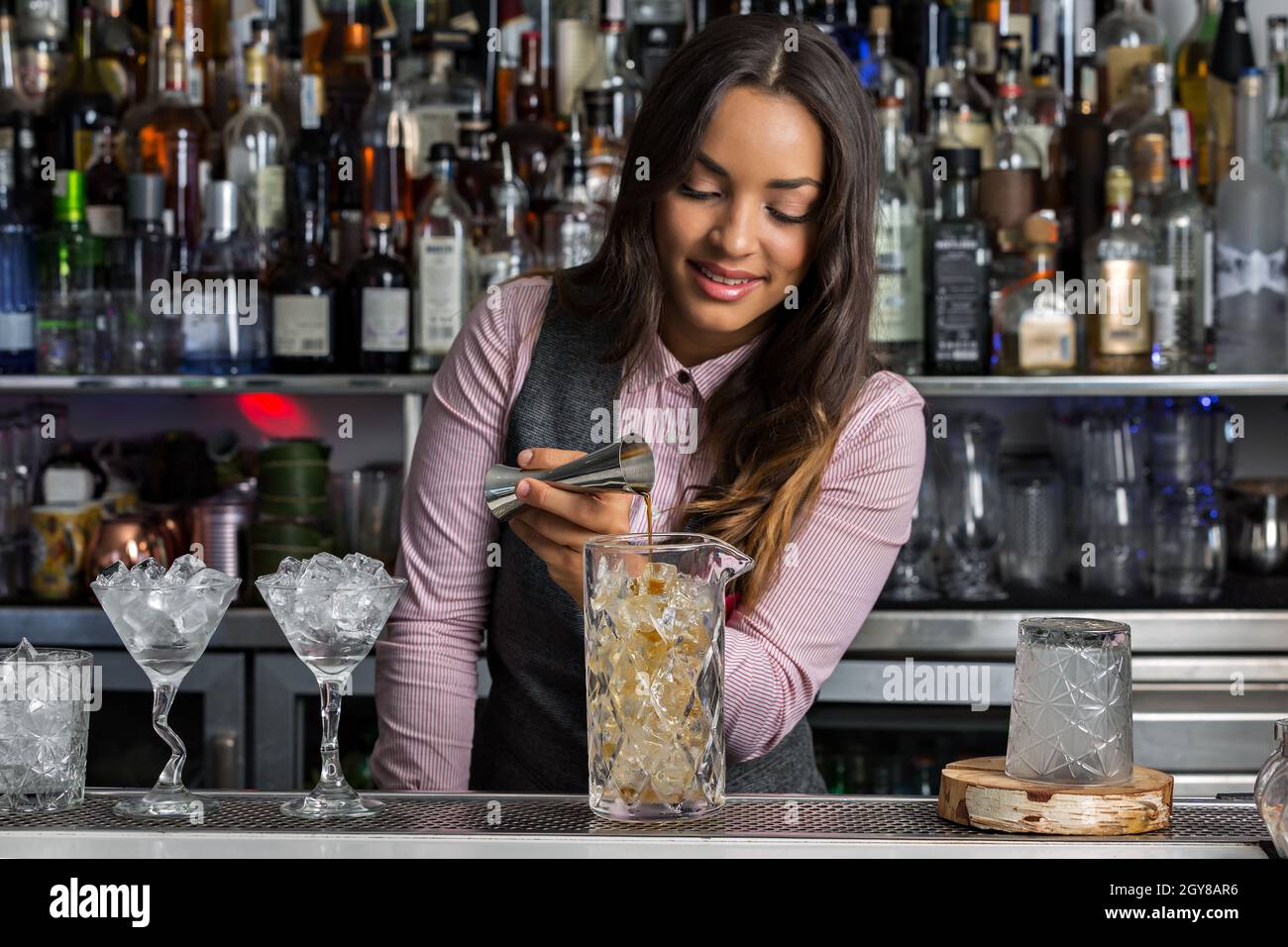 Young Hispanic female bartender adding alcohol from stainless jigger into mixing glass with ice ...