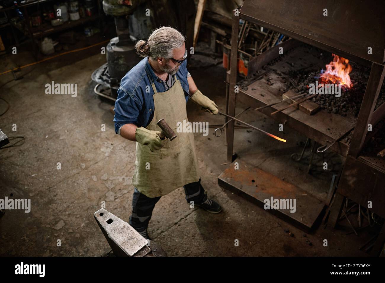 Mature blacksmith in protective wear adding coals in furnace and ...