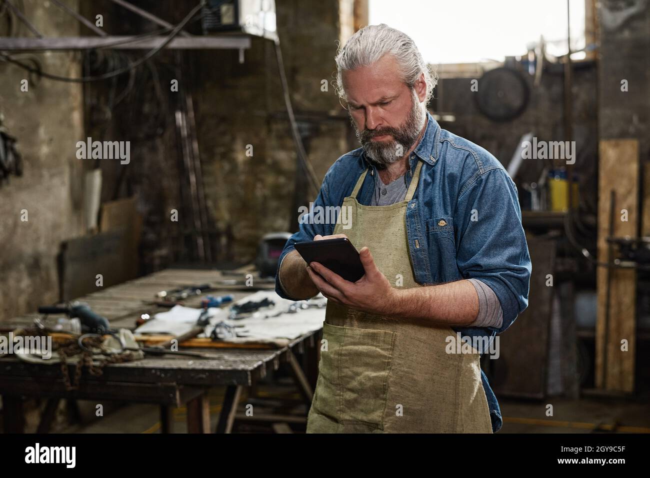 Mature carpenter in apron standing near his workplace and typing on ...