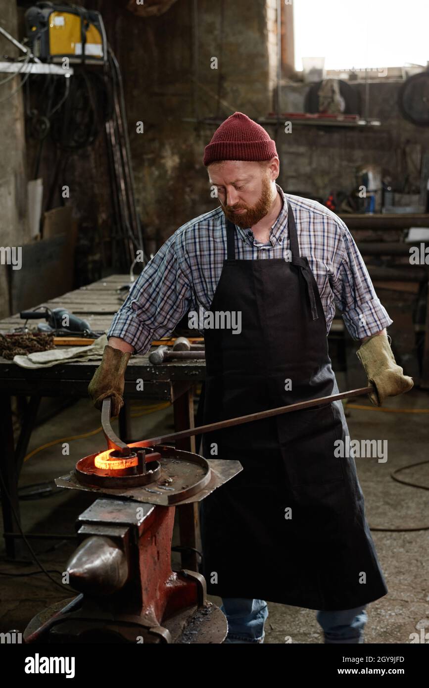 Mature bearded blacksmith forging the iron and making metallic details ...