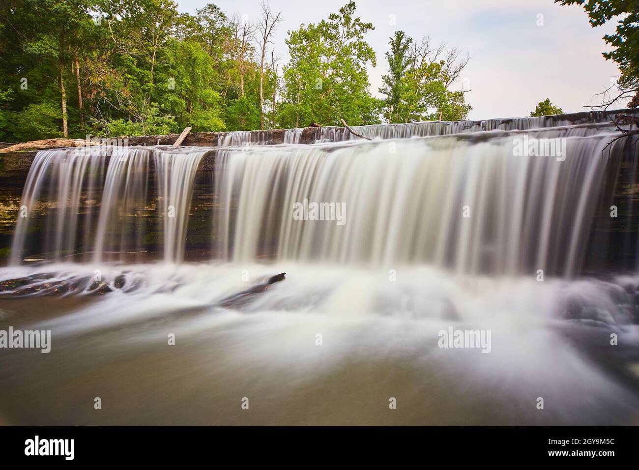 Smooth waterfall with basin below and green forest and sky above Stock ...