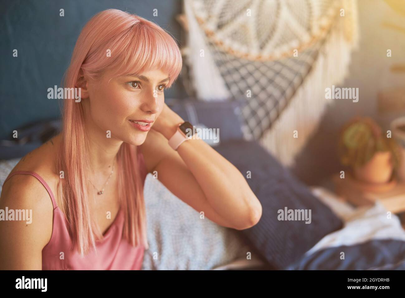 Elegant woman with pink hair and modern smartwatch sits on bed with ...