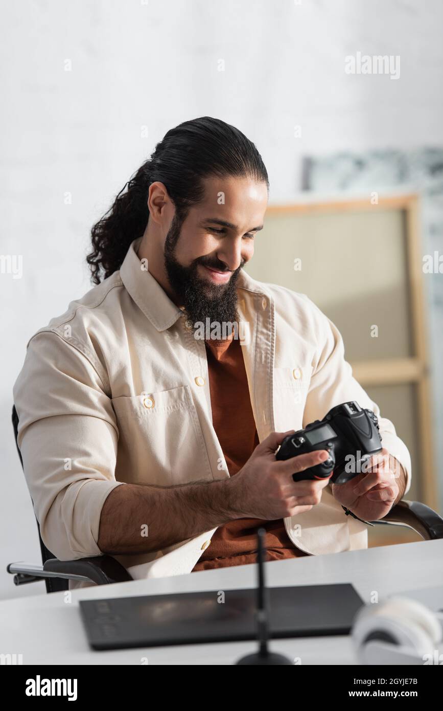 cheerful hispanic photographer looking at modern digital camera at home ...
