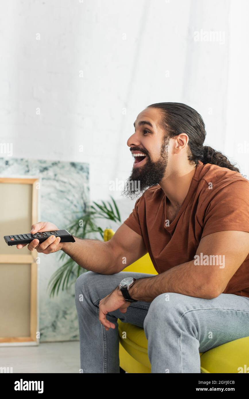 laughing latin man with remote controller watching comedy film at home ...