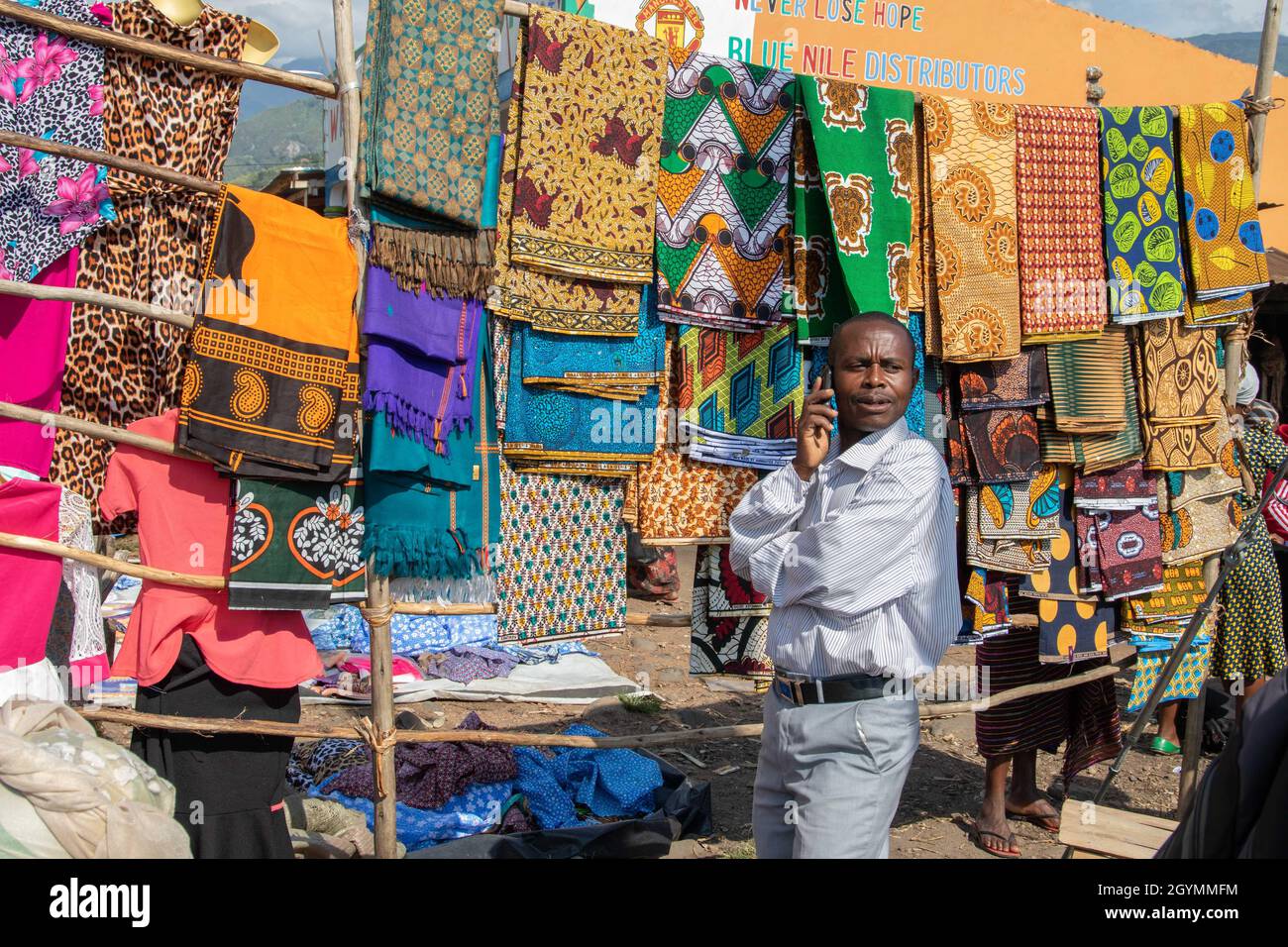 A man with his phone stands near a clothing shop on the street. Rwanda ...