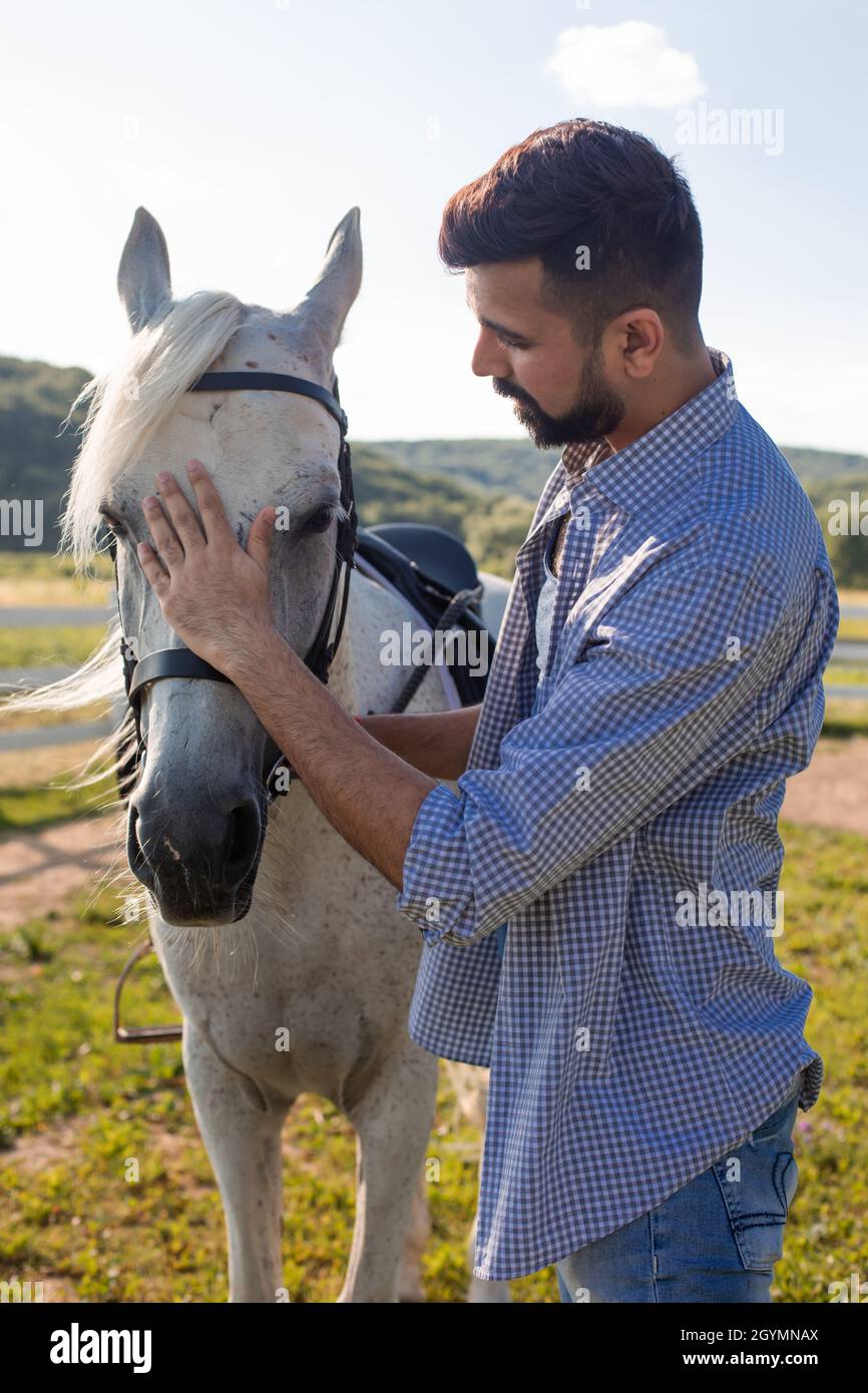 The handsome bearded man is standing next to a whte horse on a farm ...