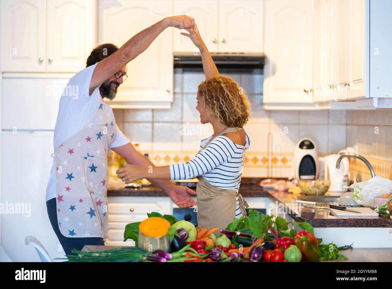 Joyful couple in apron holding hands and dancing together in domestic ...