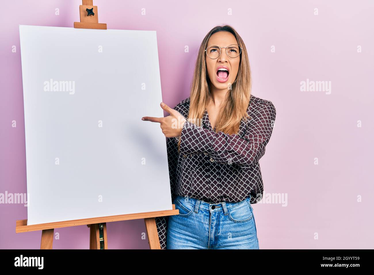 Beautiful hispanic woman pointing to painter easel stand angry and mad ...