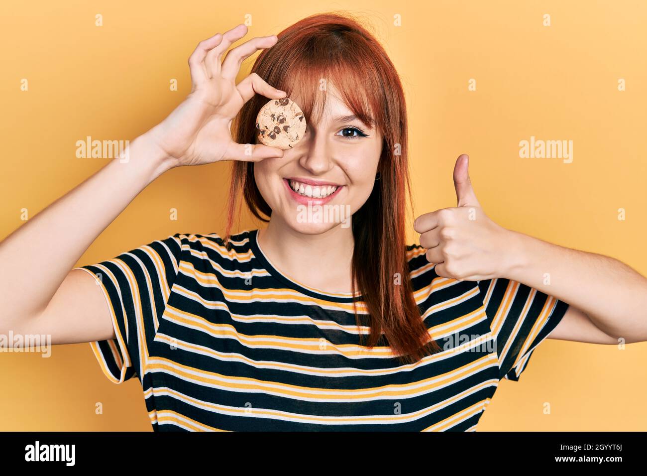 Redhead young woman holding cookie close to eye smiling happy and ...