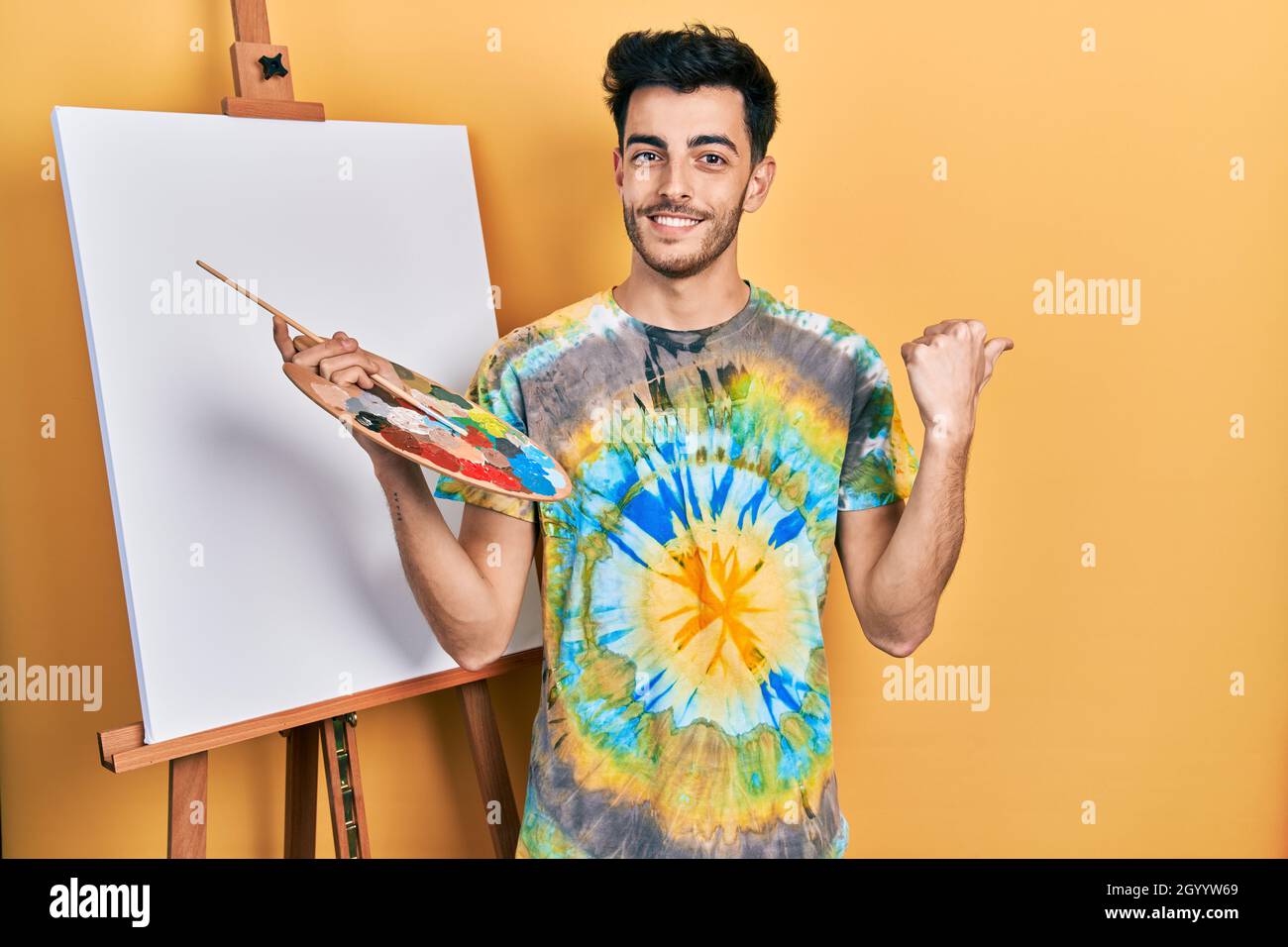 Young hispanic man standing drawing with palette by painter easel stand ...