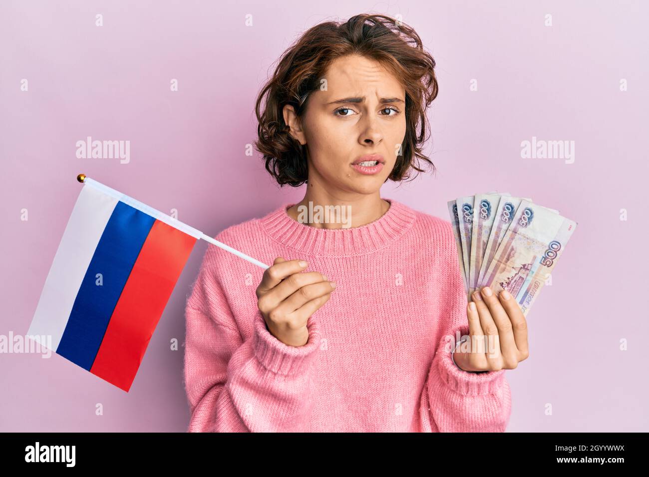 Young brunette woman holding russia flag and rubles banknotes clueless ...
