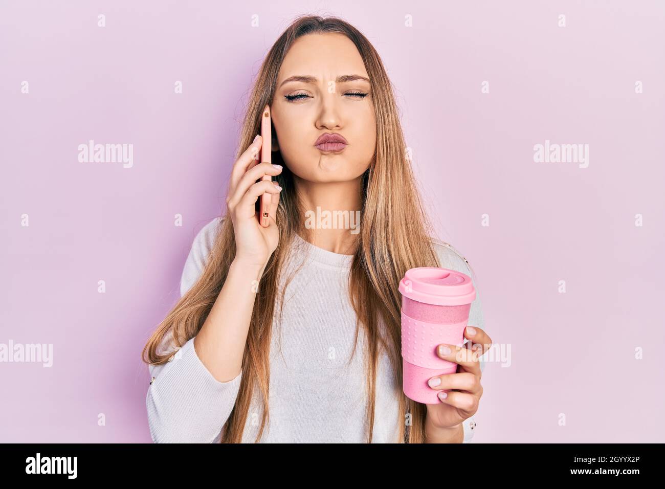 Young blonde girl using smartphone and drinking a cup of coffee puffing ...
