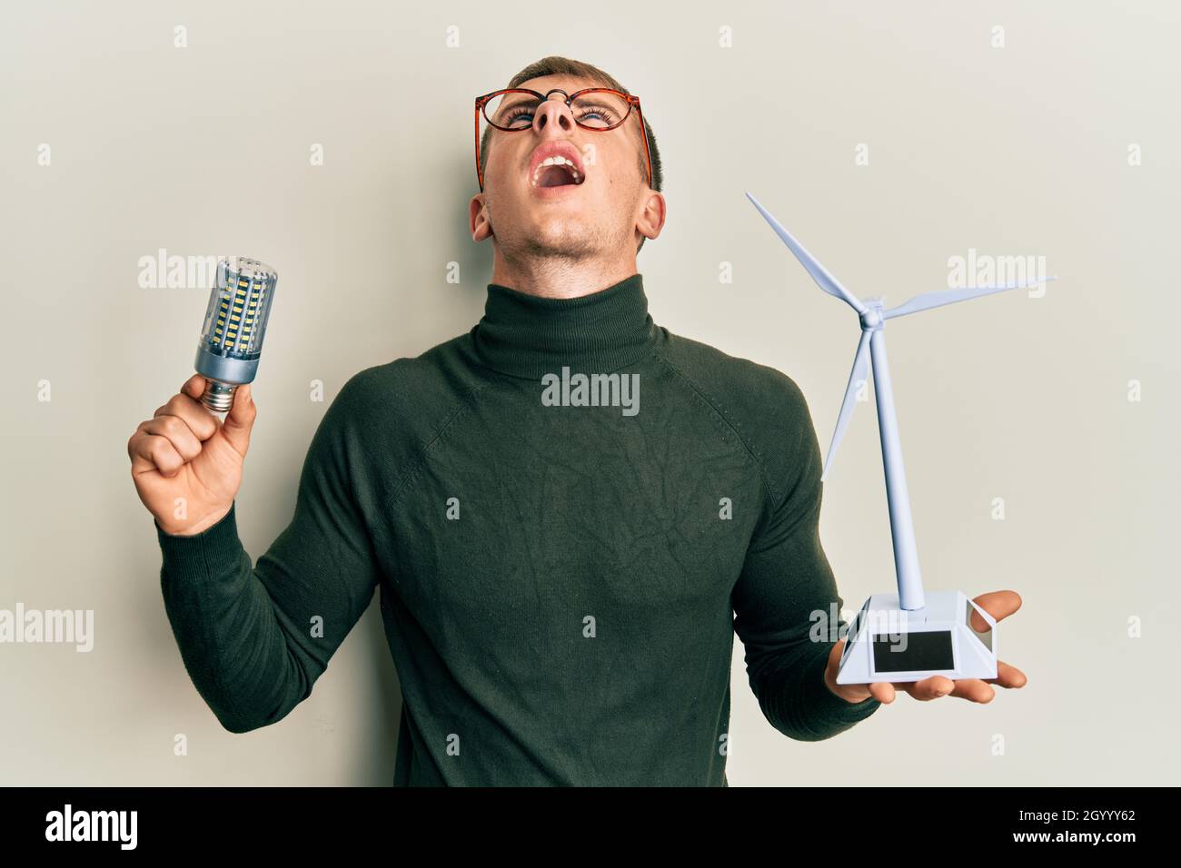 Young caucasian man holding solar windmill and led lightbulb angry and ...