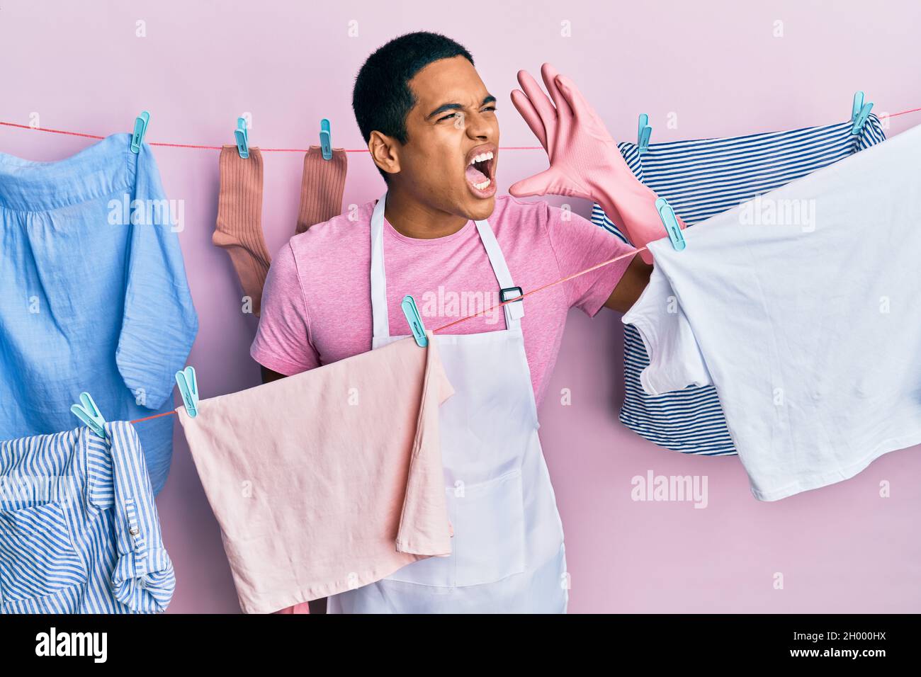 Young handsome hispanic man wearing cleaner apron holding clothes on ...