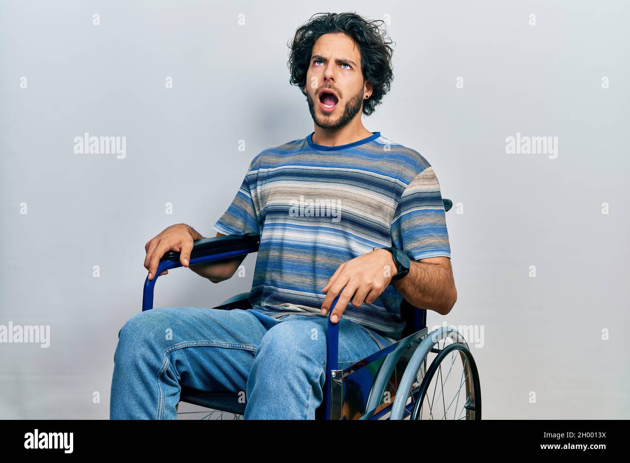 Handsome hispanic man sitting on wheelchair angry and mad screaming ...