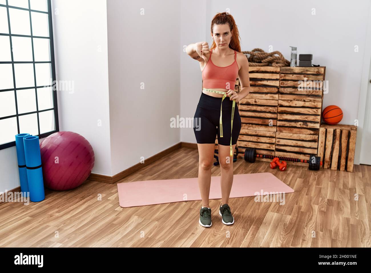 Young redhead woman using tape measure measuring waist at the gym with ...