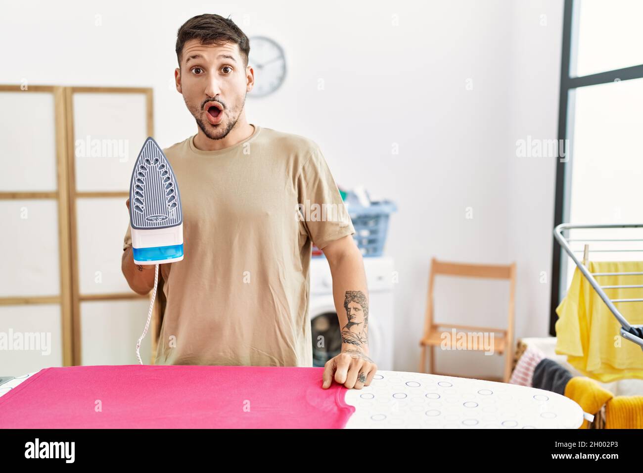 Young handsome man ironing clothes at home scared and amazed with open ...