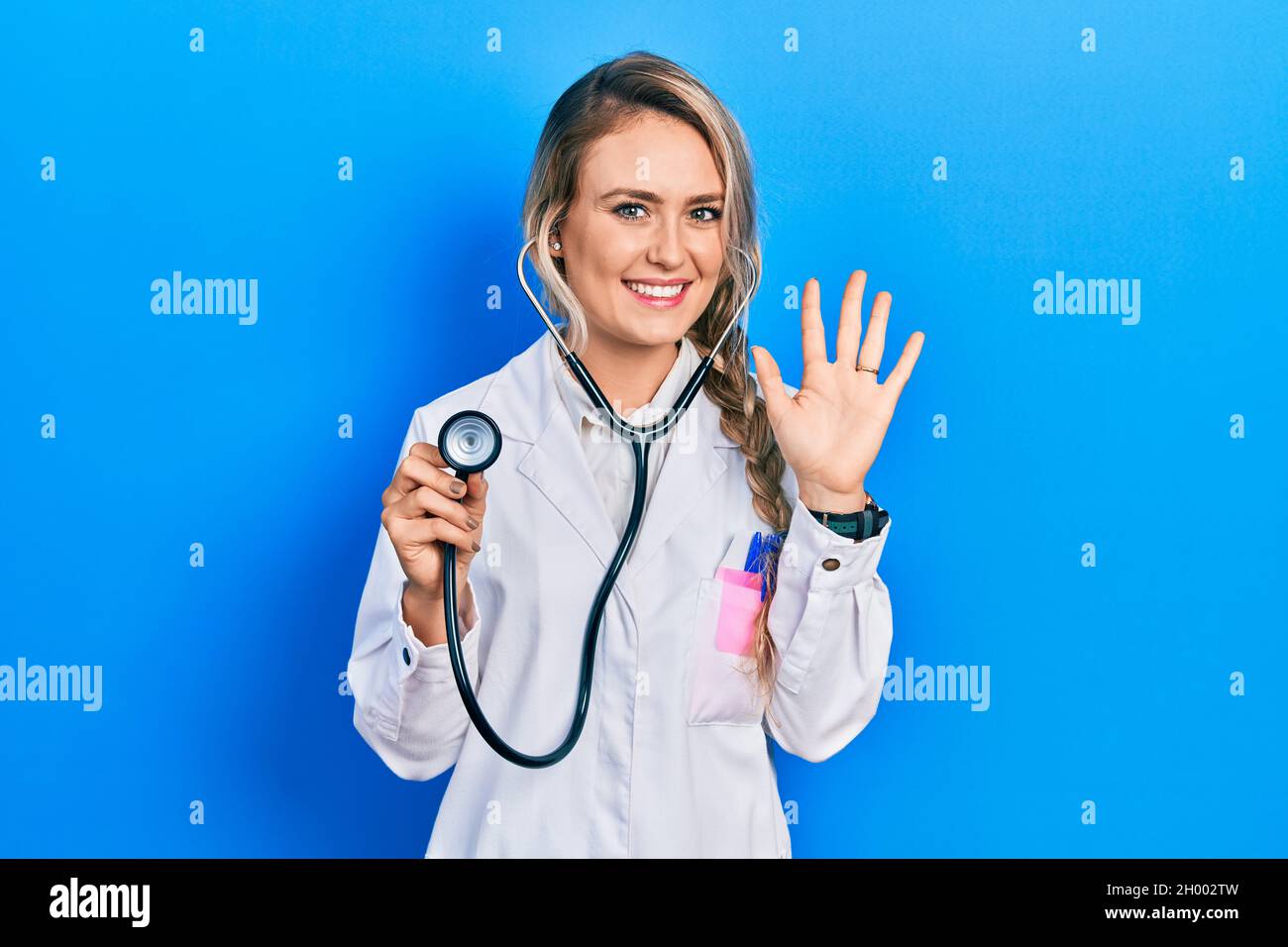 Beautiful young blonde doctor woman holding stethoscope waiving saying ...