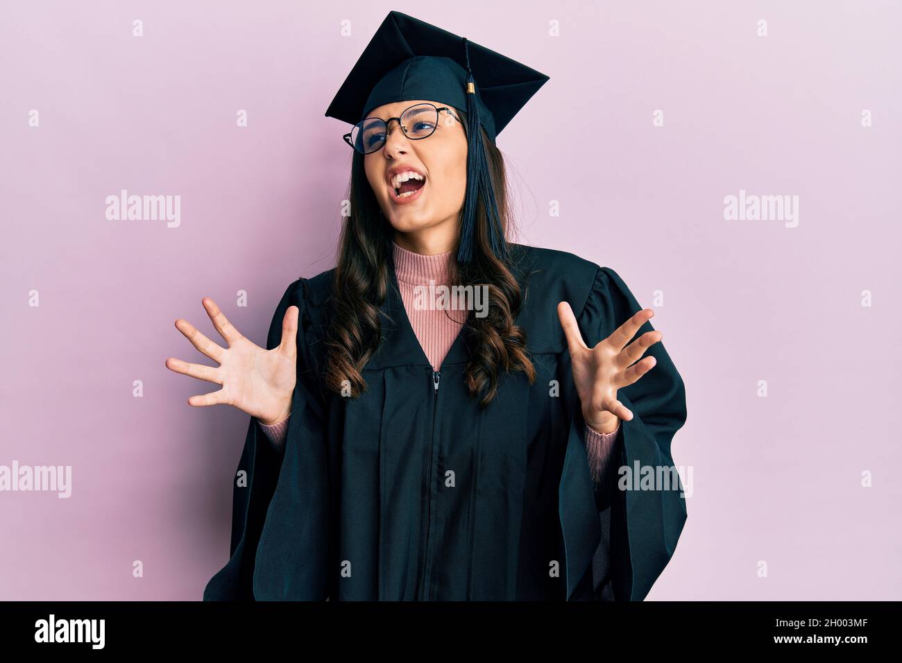 Young hispanic woman wearing graduation cap and ceremony robe crazy and ...