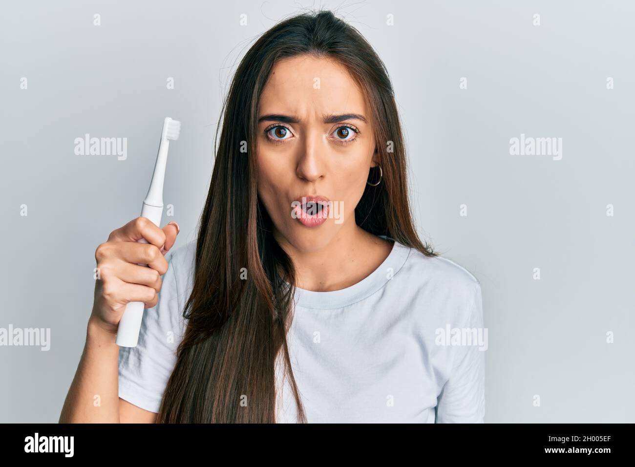Young hispanic girl holding electric toothbrush scared and amazed with ...