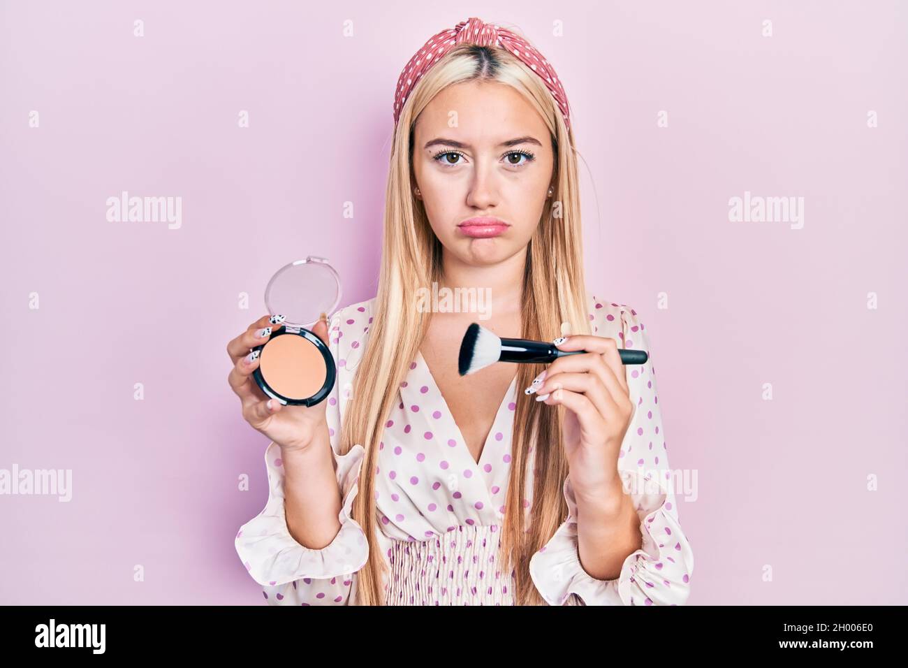 Young blonde girl holding makeup bronzer and brush depressed and worry ...