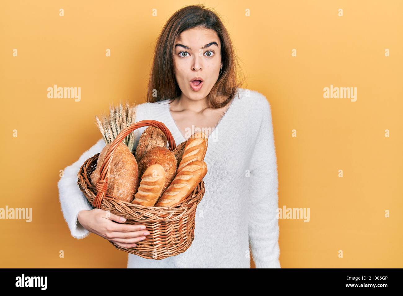 Young caucasian girl holding wicker basket with bread scared and amazed ...