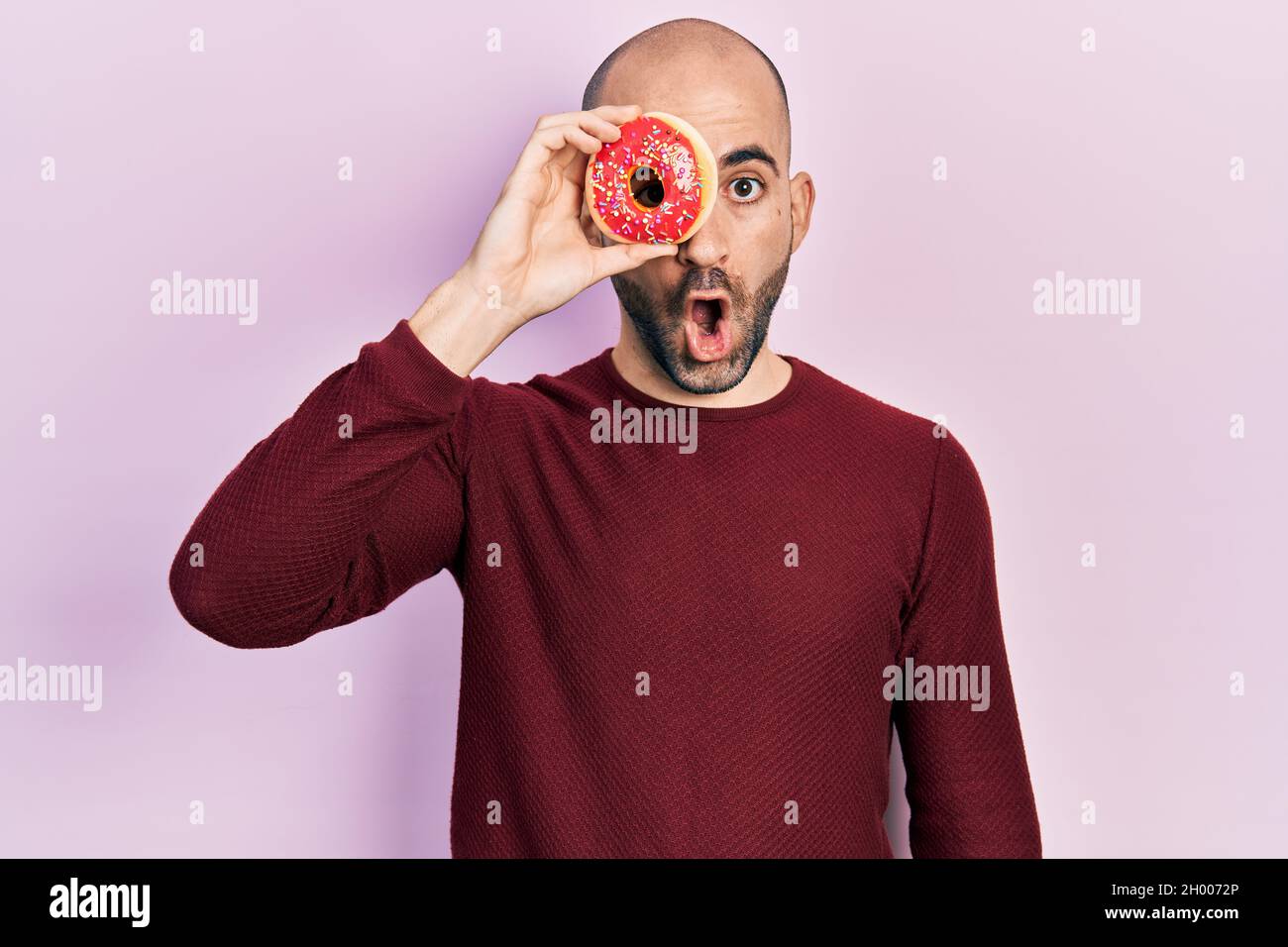 Young bald man holding tasty colorful doughnut on eye scared and amazed ...