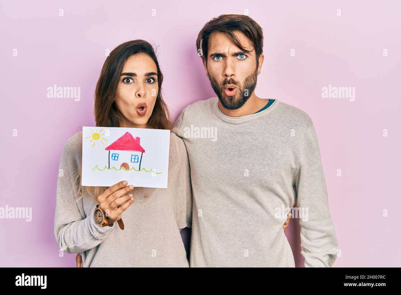 Young hispanic couple holding house draw scared and amazed with open ...