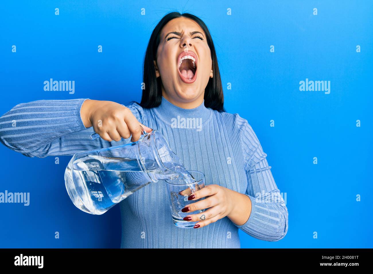 Beautiful brunette woman pouring water in glass angry and mad screaming ...