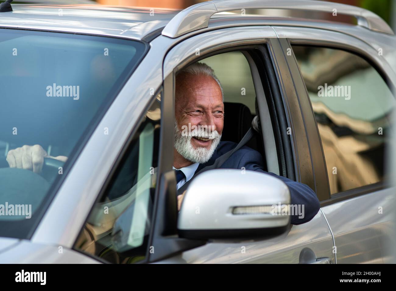 Portrait of happy senior man in business suit driving car with elbow on ...