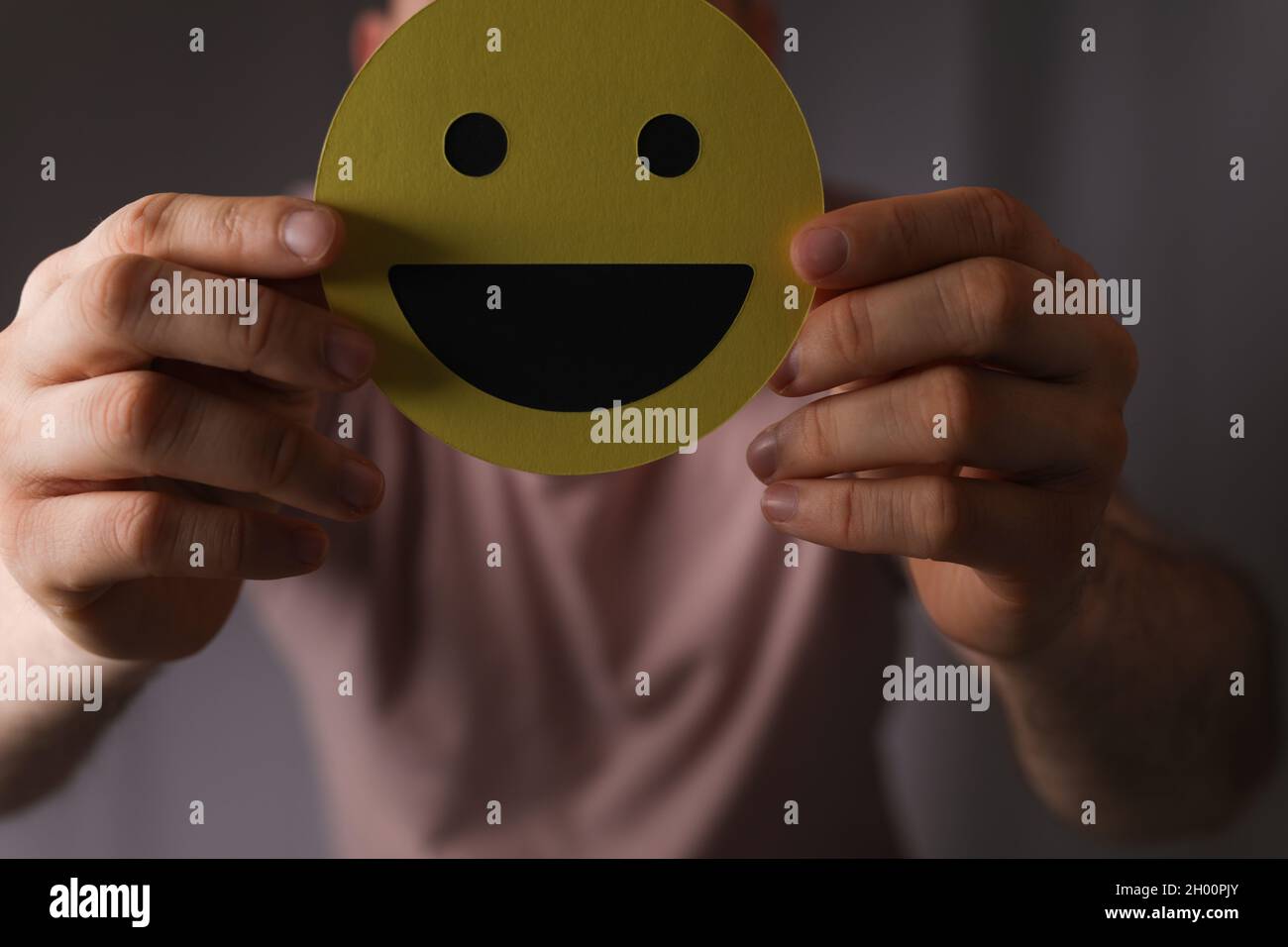 A closeup of a person holding a yellow smiley face in front of his face ...