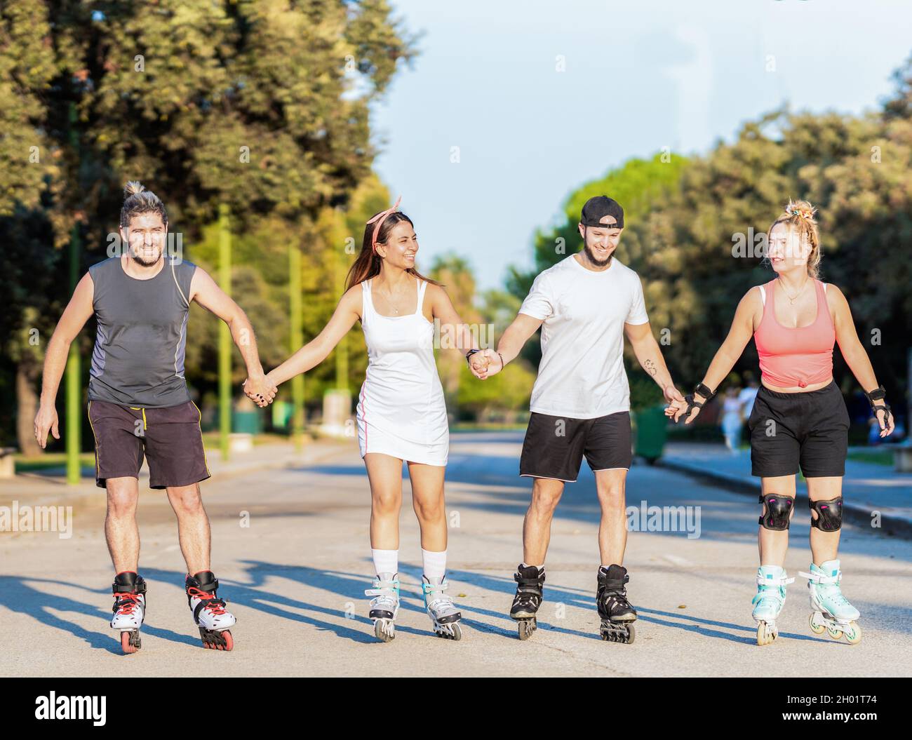 Friends skating on inline skates holding hands on a street with trees ...