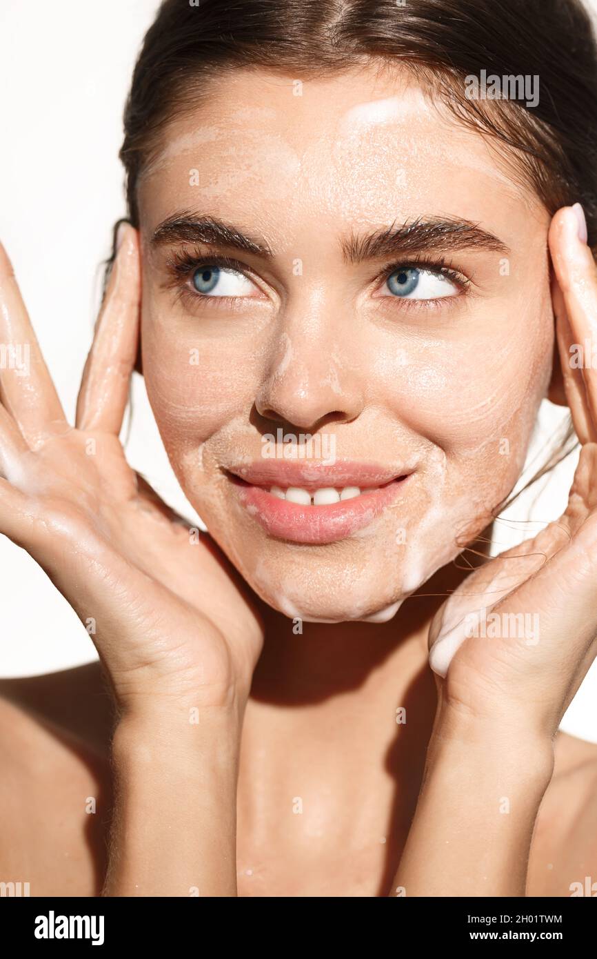 Vertical portrait of young woman washing her face with cleansing foam gel, smiling happy ...