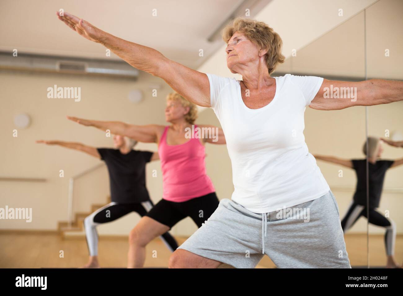 Senior woman performing lunging asana Virabhadrasana during group yoga ...