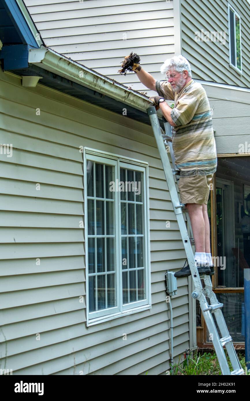 An older man carefully stands on a ladder and cleans leaves and debris ...