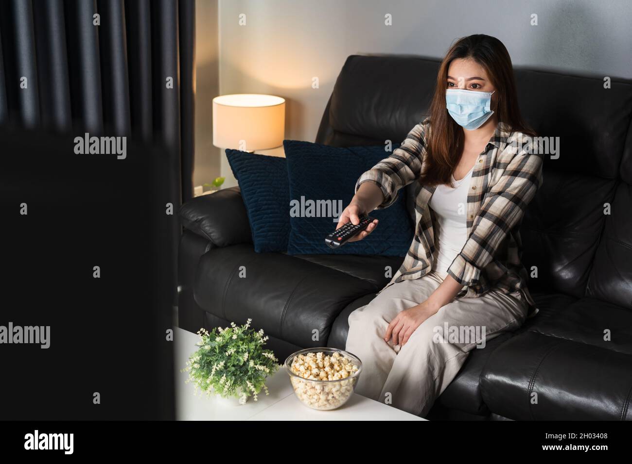 young woman watching TV on sofa and wearing medical mask to protect ...