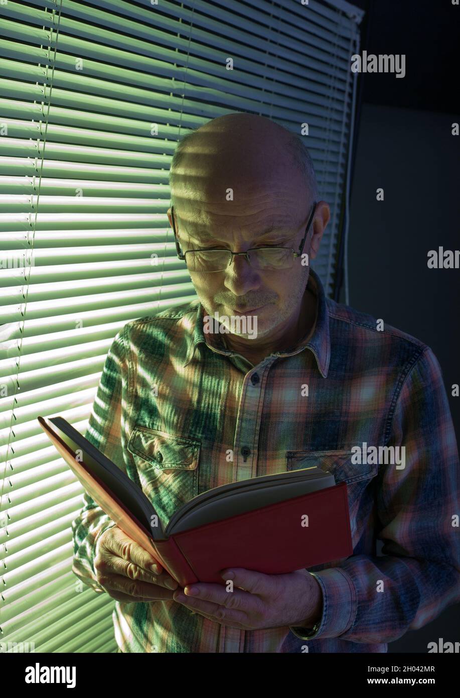 Senior man reading book beside window with blinds Stock Photo - Alamy