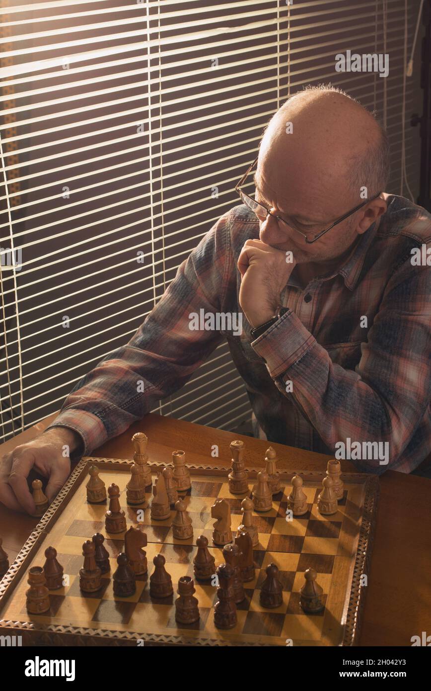 Old man sitting at table beside window with blinds and playing chess ...
