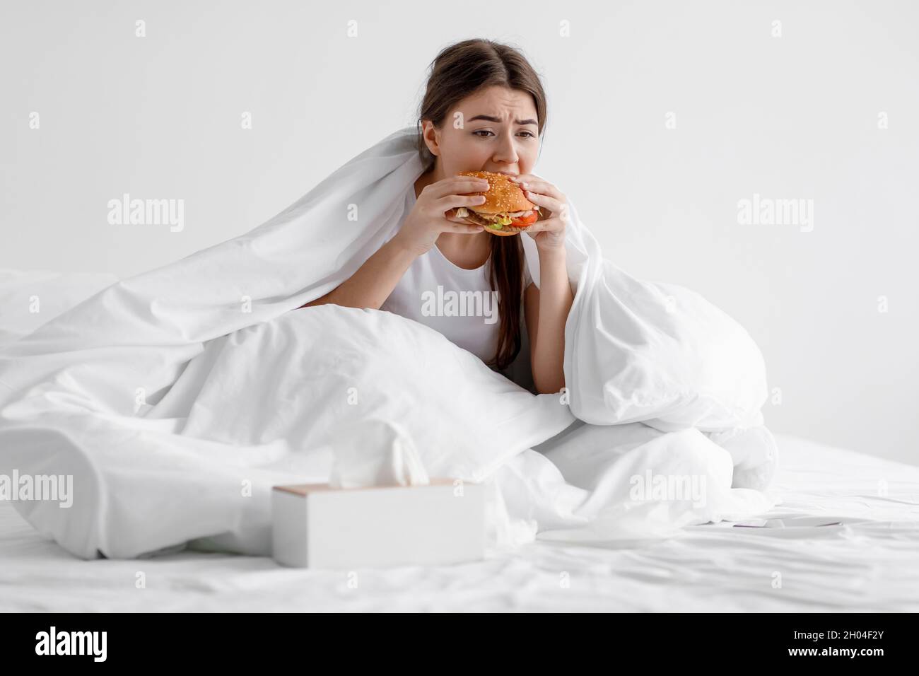 Upset hungry european millennial woman eating big burger, sits on white ...
