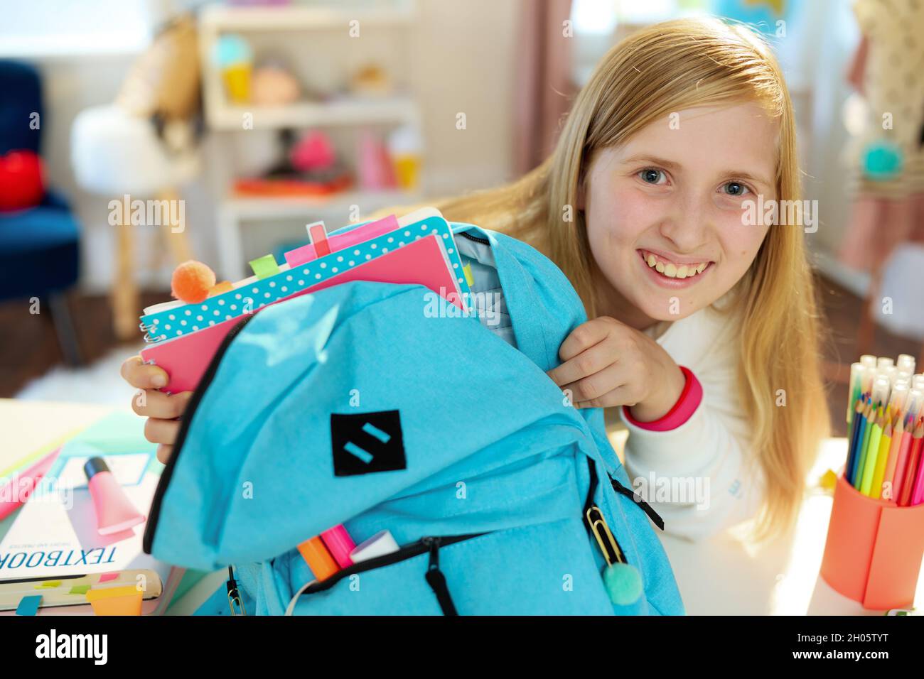 smiling modern girl with workbooks and blue backpack packing for school ...