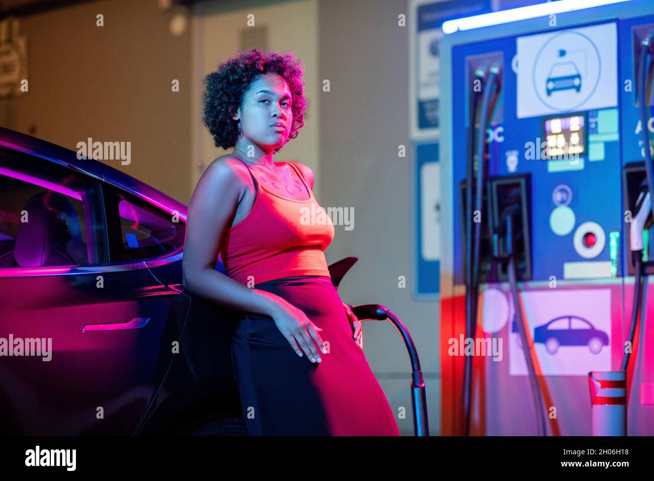 Young female standing by electric car against fueling pile during ...