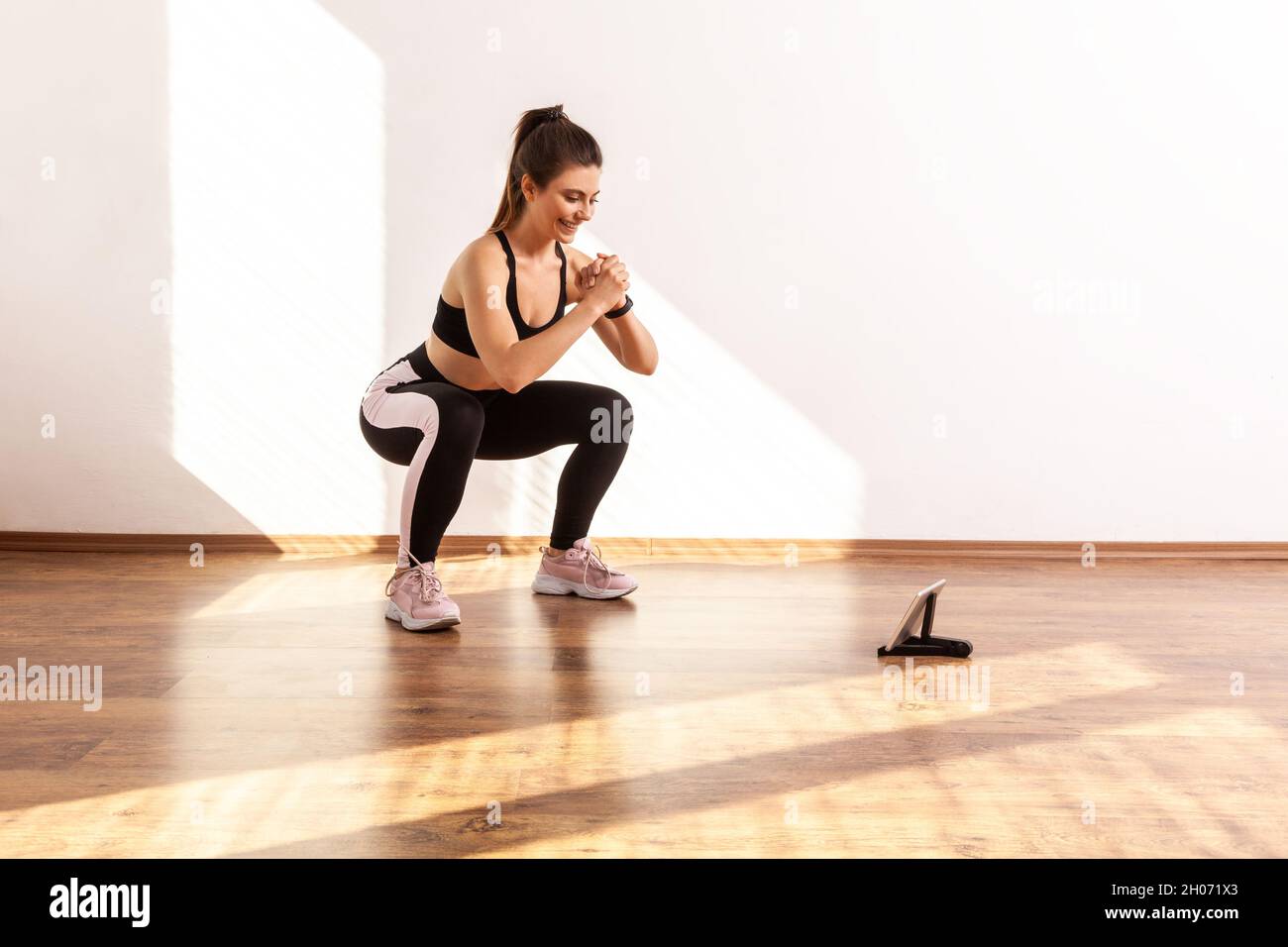 Athletic female doing squats while looking online fitness class ...