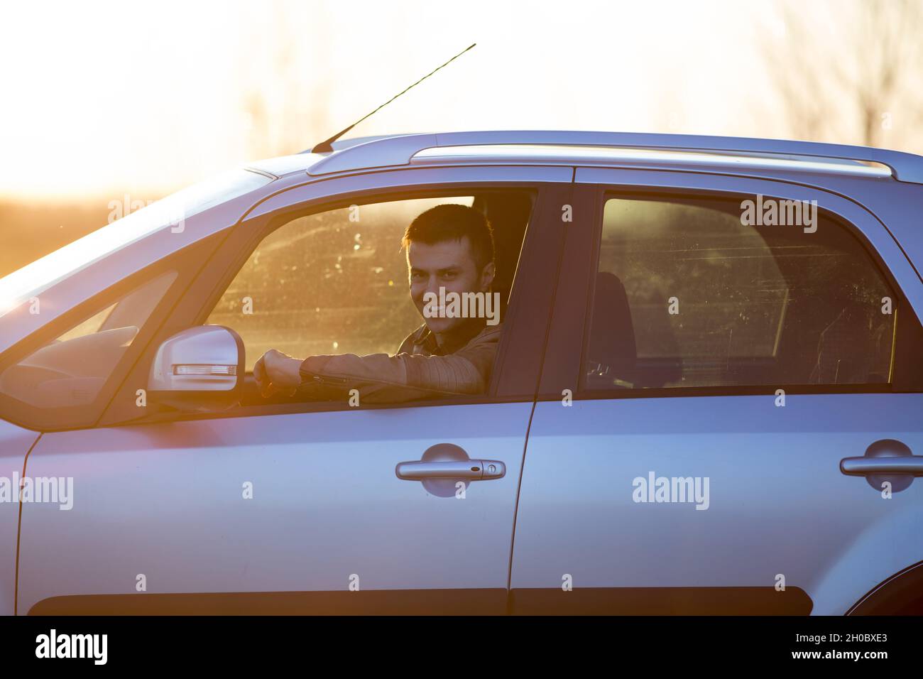 Handsome young man driving car and looking through window at sunset ...